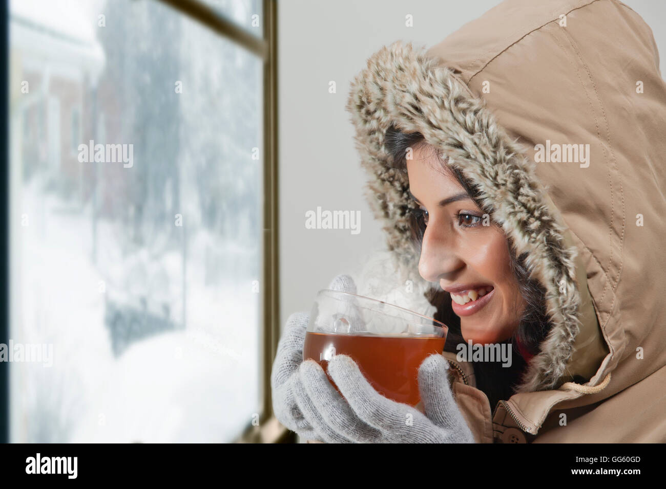 Smiling young woman looking through window and drinking tea Stock Photo ...