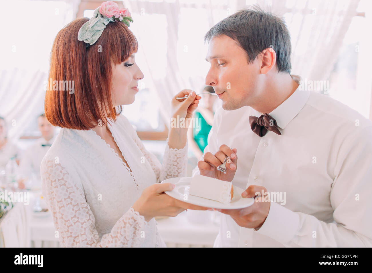 Beautiful young bride feeding wedding cake to groom in light restaurant ...