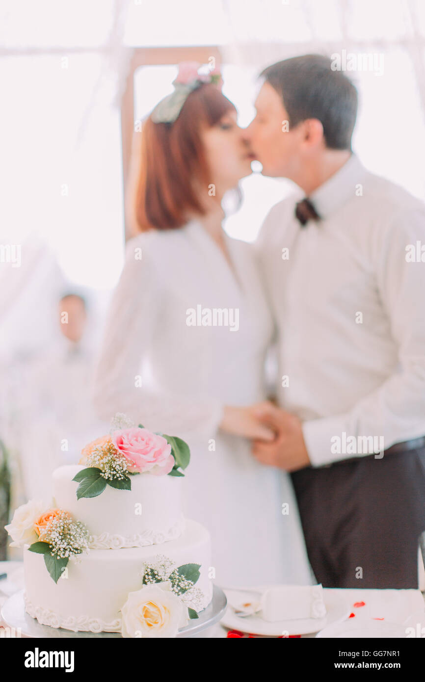 Kiss of the newlyweds next to the white wedding cake decorated with ...