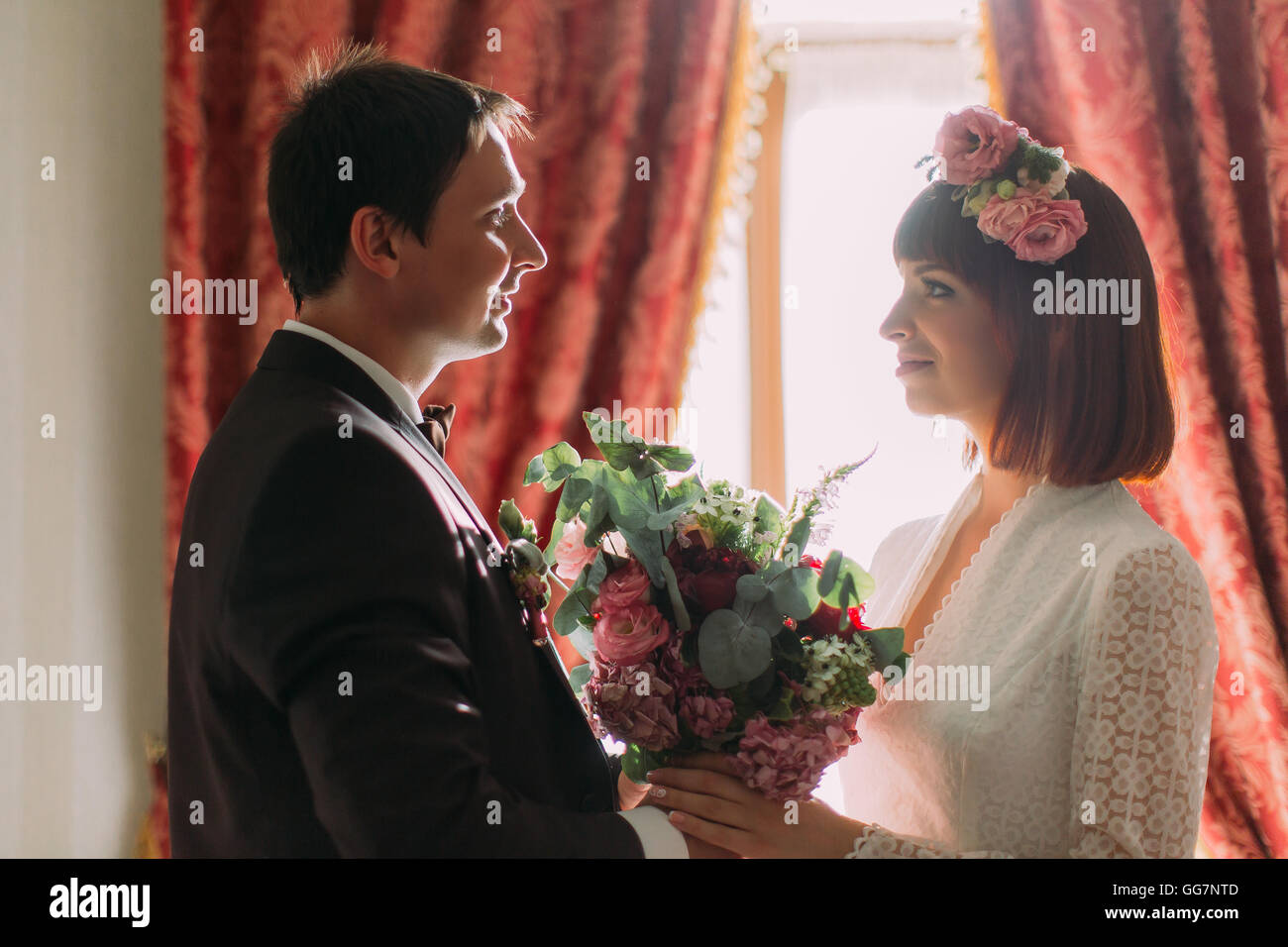 Happy luxury bride and groom looking at each other standing near window ...