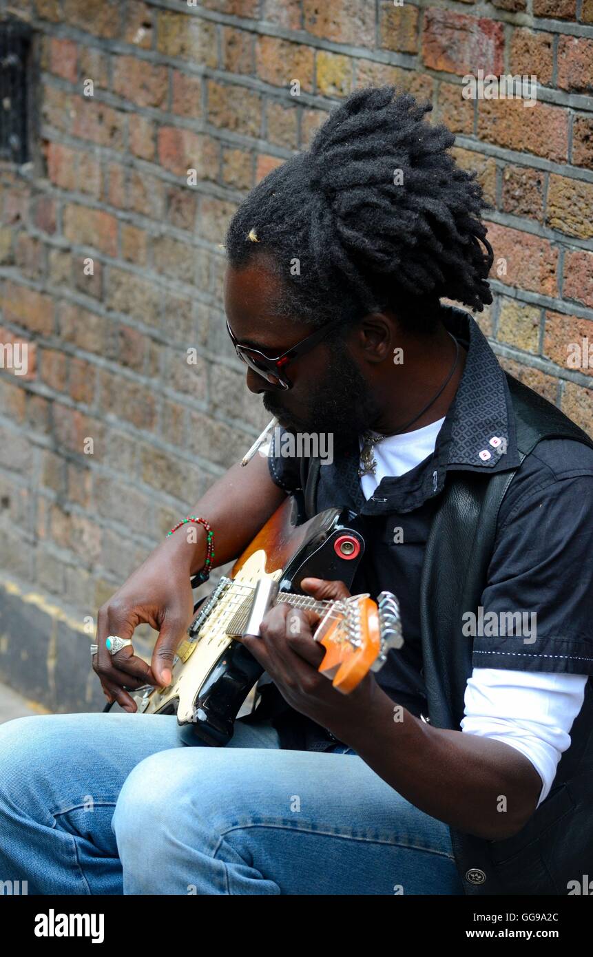 Black street musician with dreadlocks sits against wall plays guitar ...