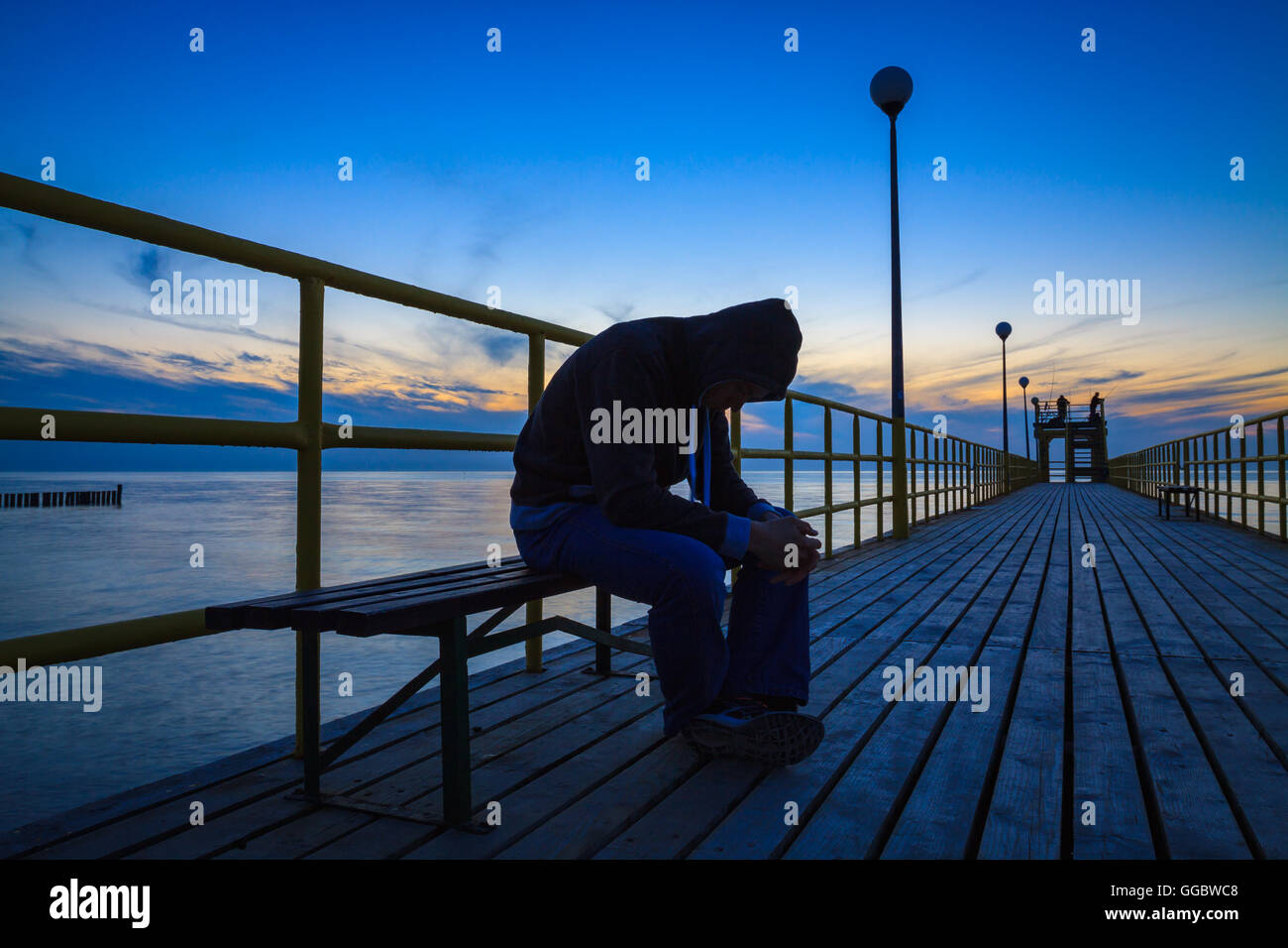 The sad guy in hood, sitting on bench at wooden pier. Baltic sea in the ...