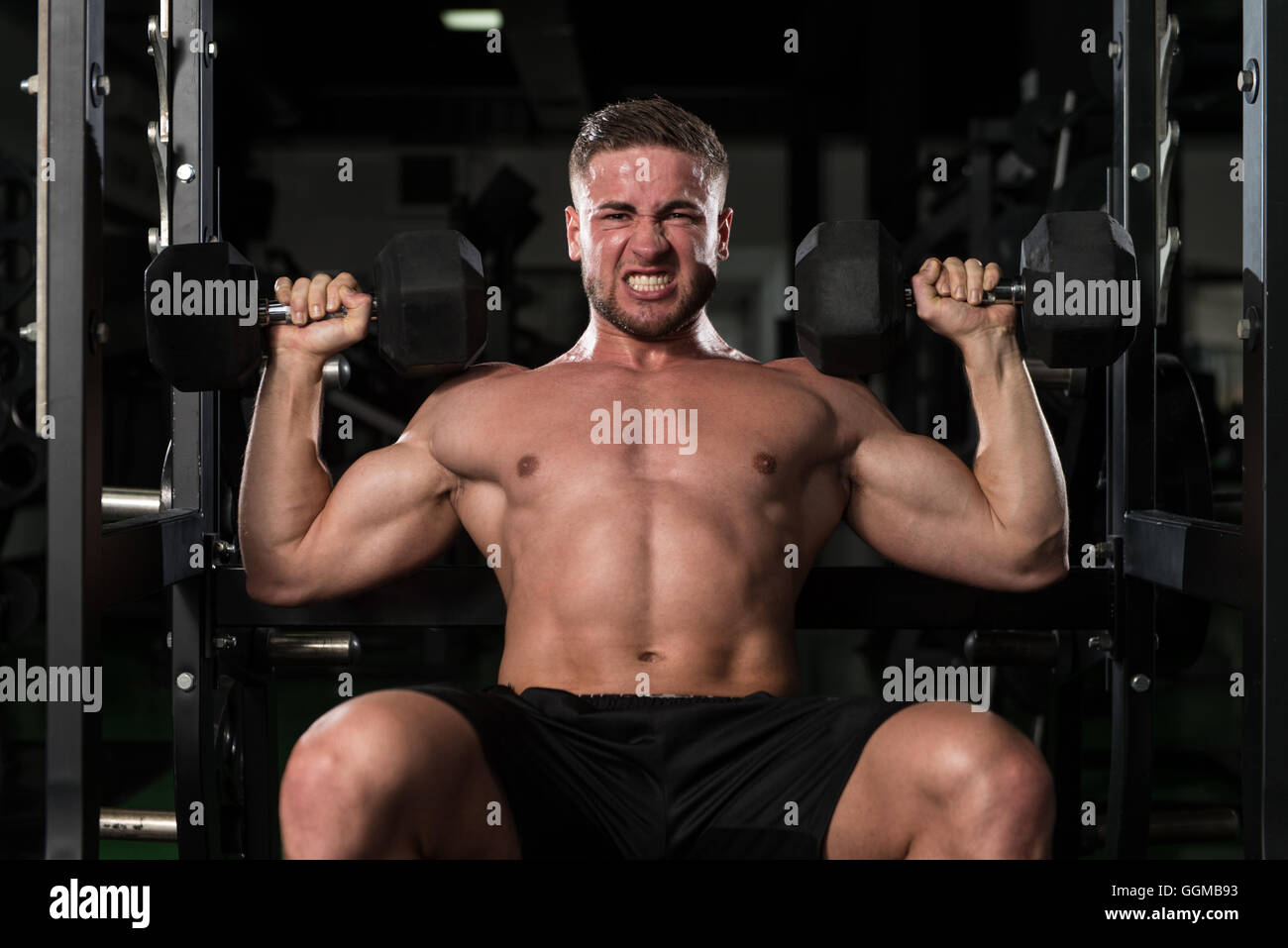 Young Strong Man In The Gym And Exercising Shoulders With Dumbbells ...