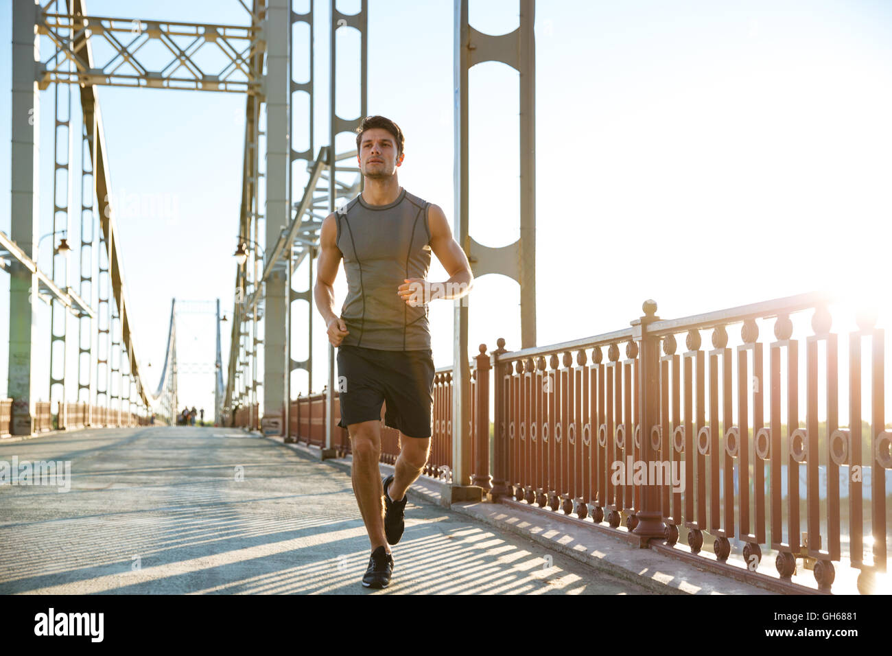 Attractive fit man running fast along big modern bridge at sunset light ...