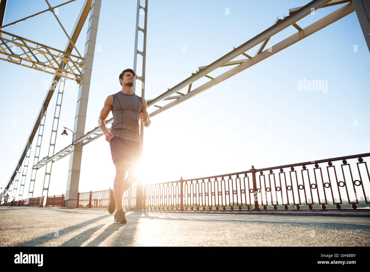 Handsome fit man running fast along big modern bridge at sunset light ...