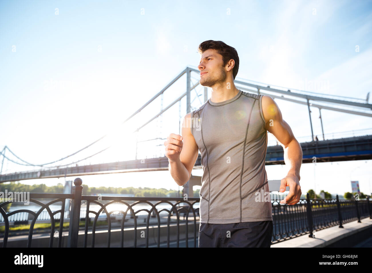 Handsome sports man running along big modern bridge at sunset light ...