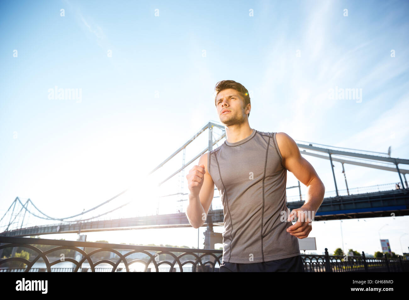 Handsome sports man running along big modern bridge at sunset light ...