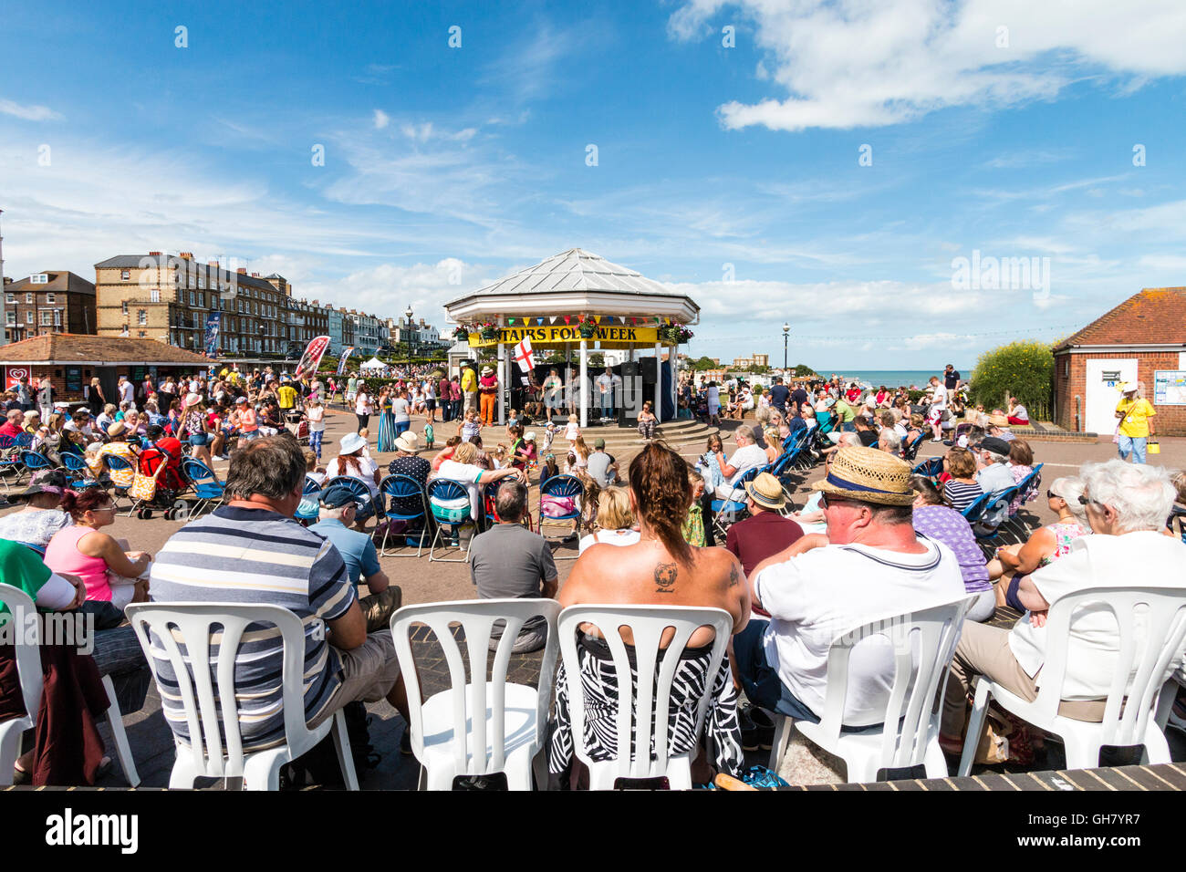 Audience of people sitting around deck and bandstand in bright sunshine ...