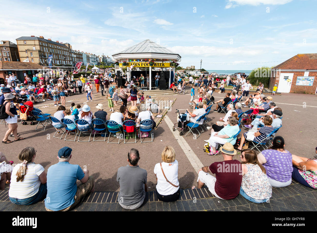 Audience of people sitting around deck and bandstand in bright sunshine ...