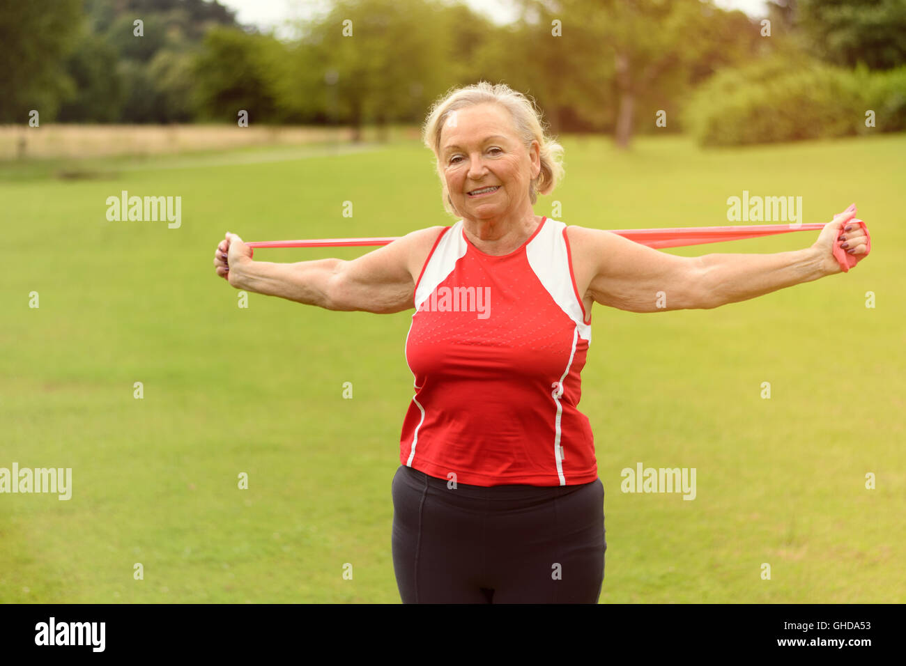 Fit senior woman in red sleeveless shirt using elastic resistance bands ...