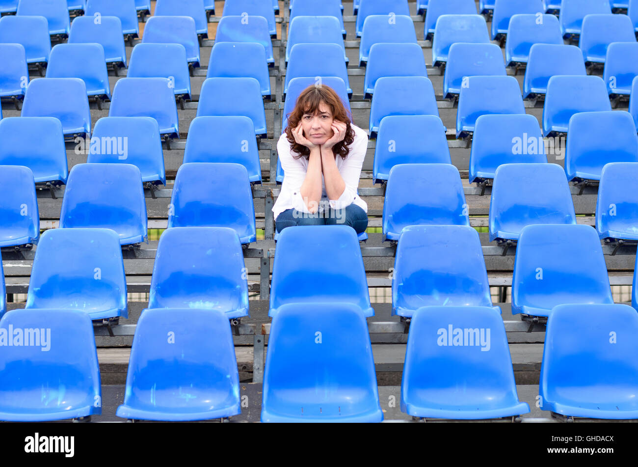 Glum woman sitting in empty rows of blue spectator seating in an ...
