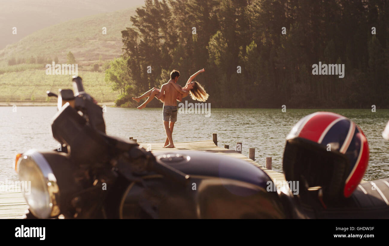 Young man carrying exuberant woman on lakeside dock behind motorcycle ...