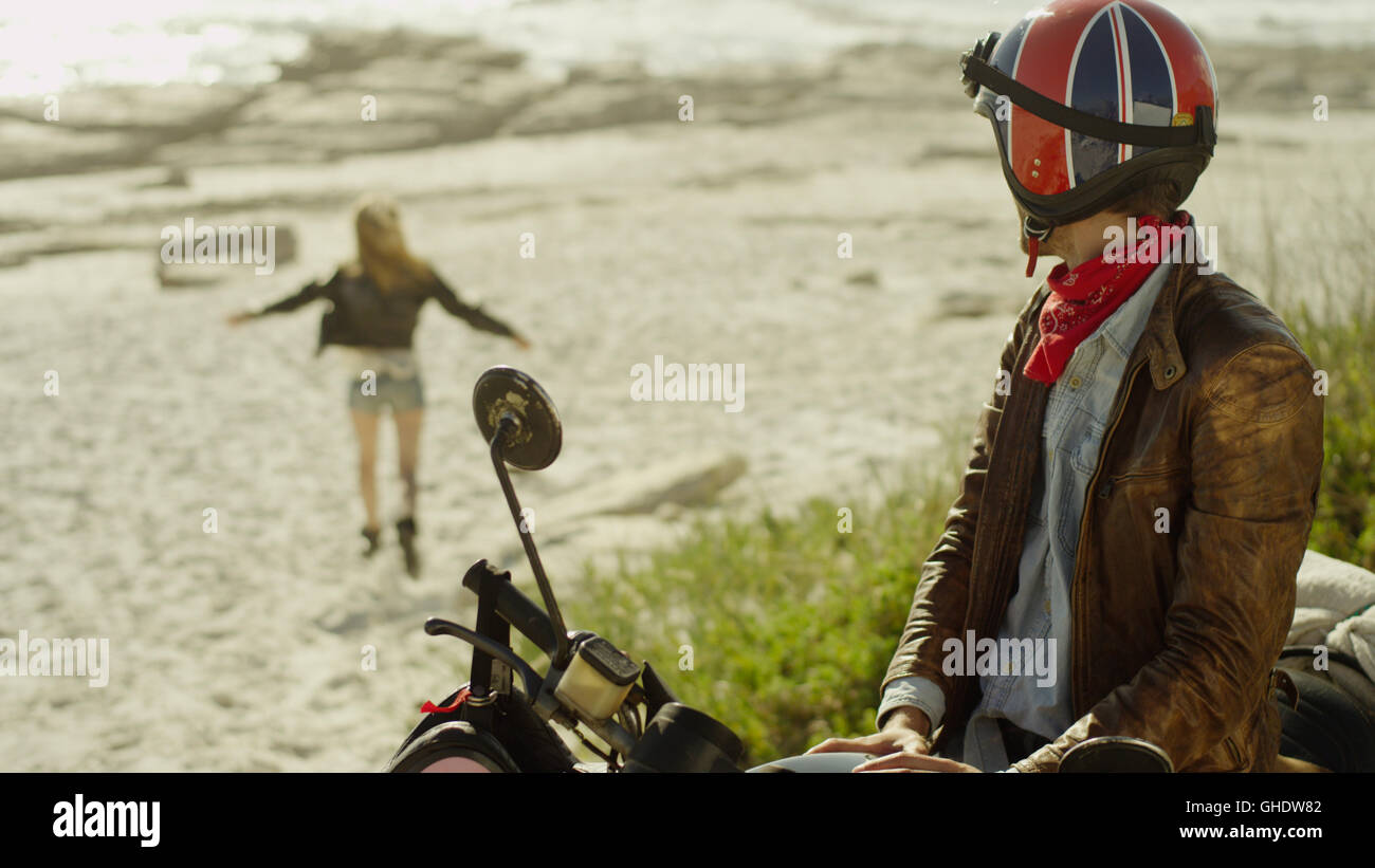 Young man on motorcycle watching woman run onto beach Stock Photo - Alamy