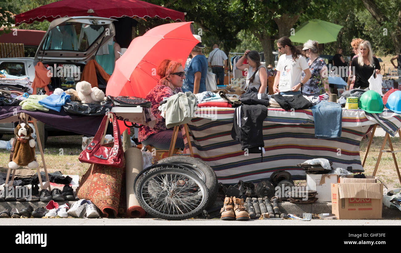 A seller at a car boot sale vide-grenier shelters under a red umbrella ...