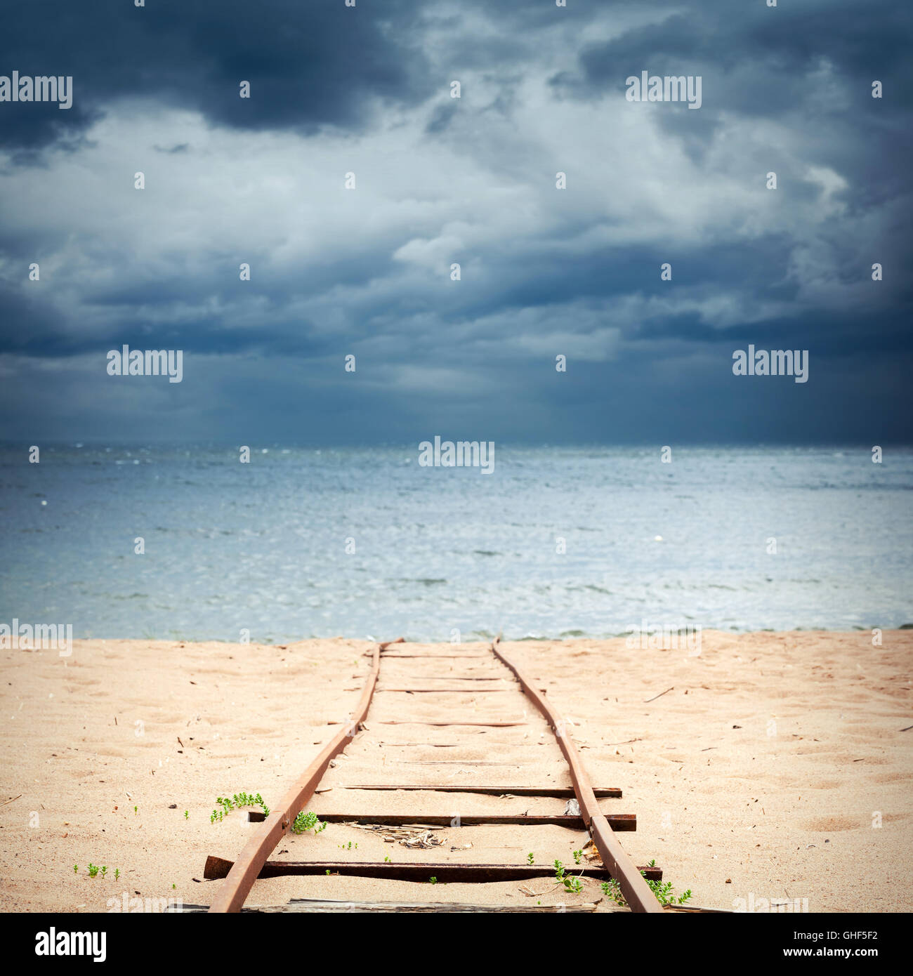 Old rusted railway goes over sandy beach to sea water under dark ...