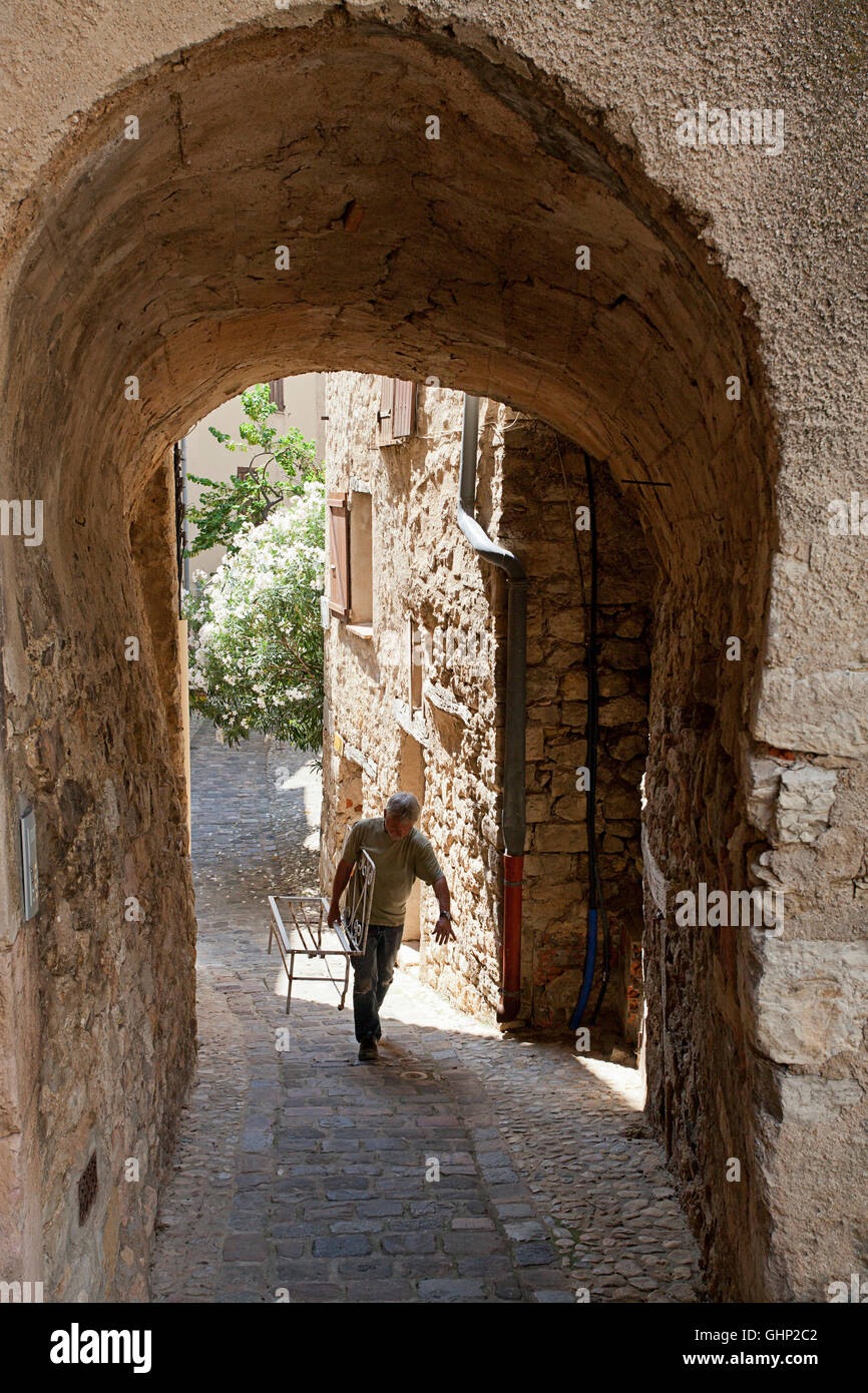 An elderly man carries a metal bench through an old French archway ...