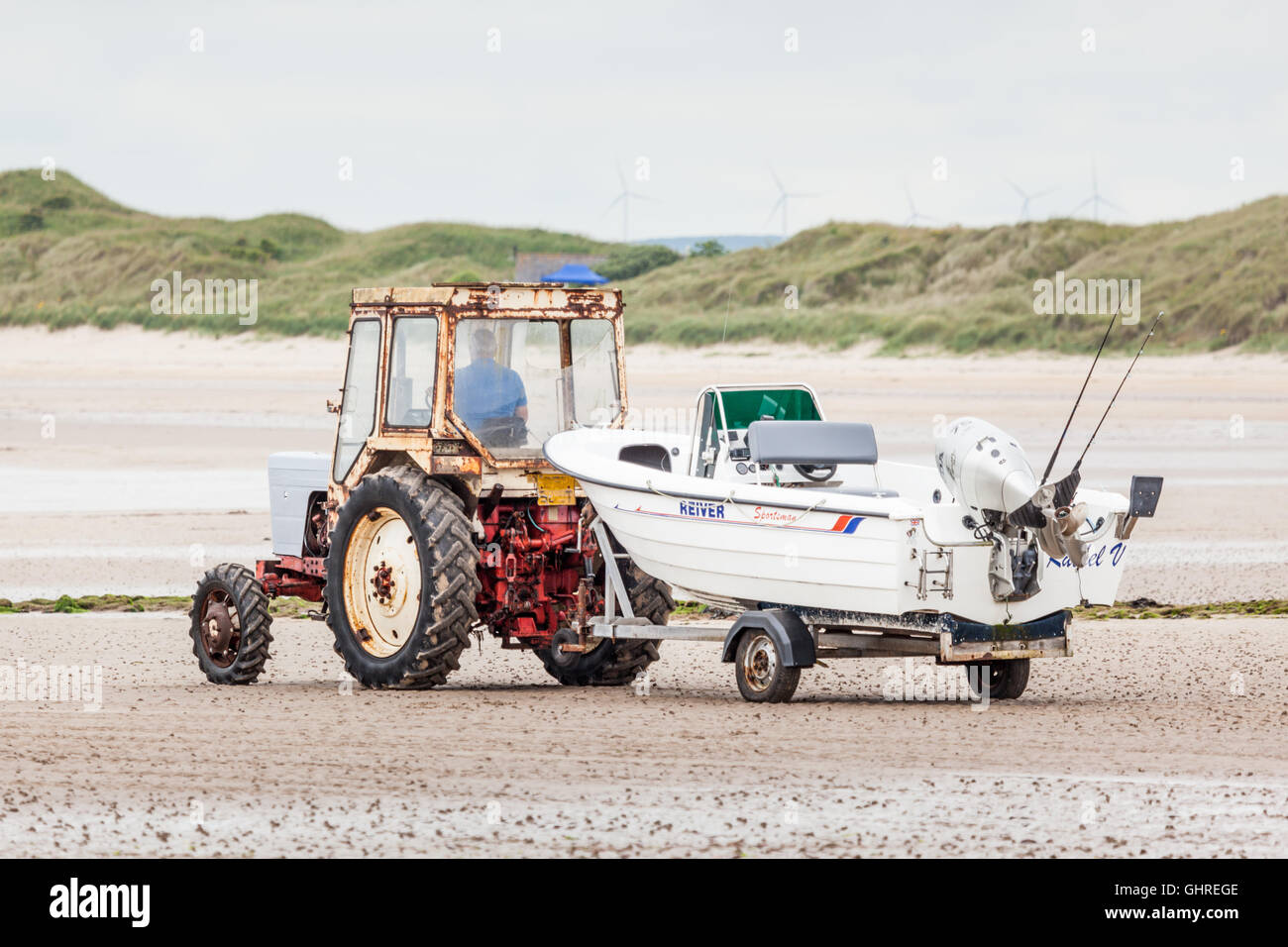 An old tractor pulling a speed boat across the beach to the sea at ...