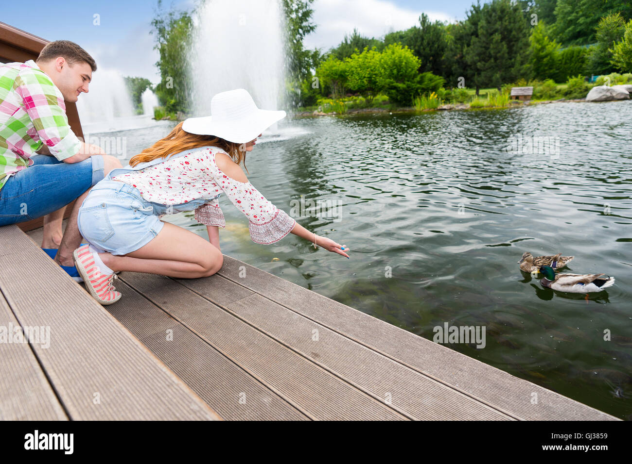 Young woman feeding ducks in a pond with her boyfriend kneeling down on ...