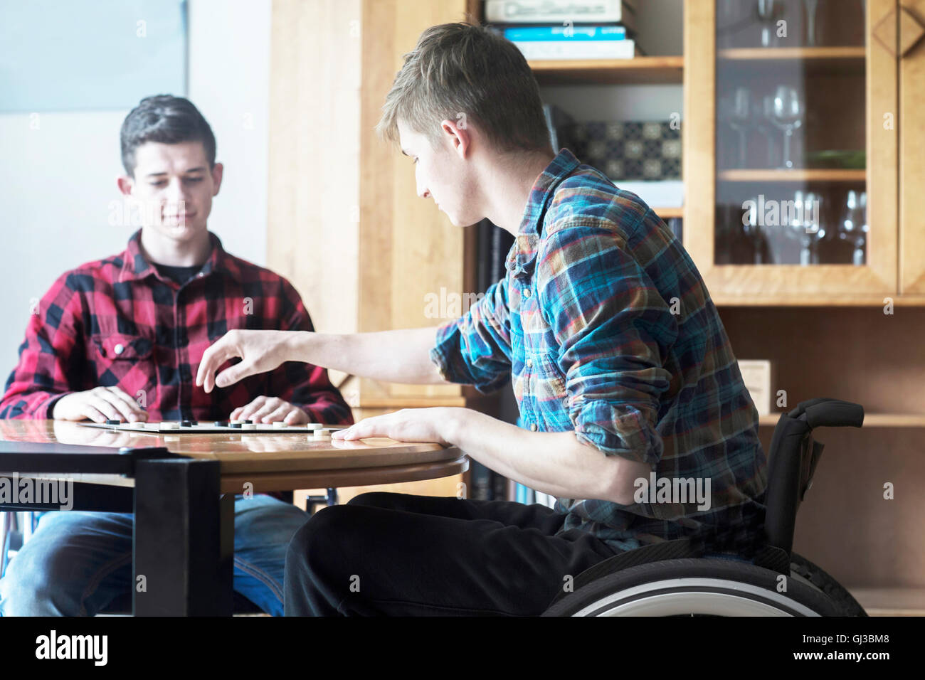 Young man using wheelchair playing draughts with friend in kitchen ...