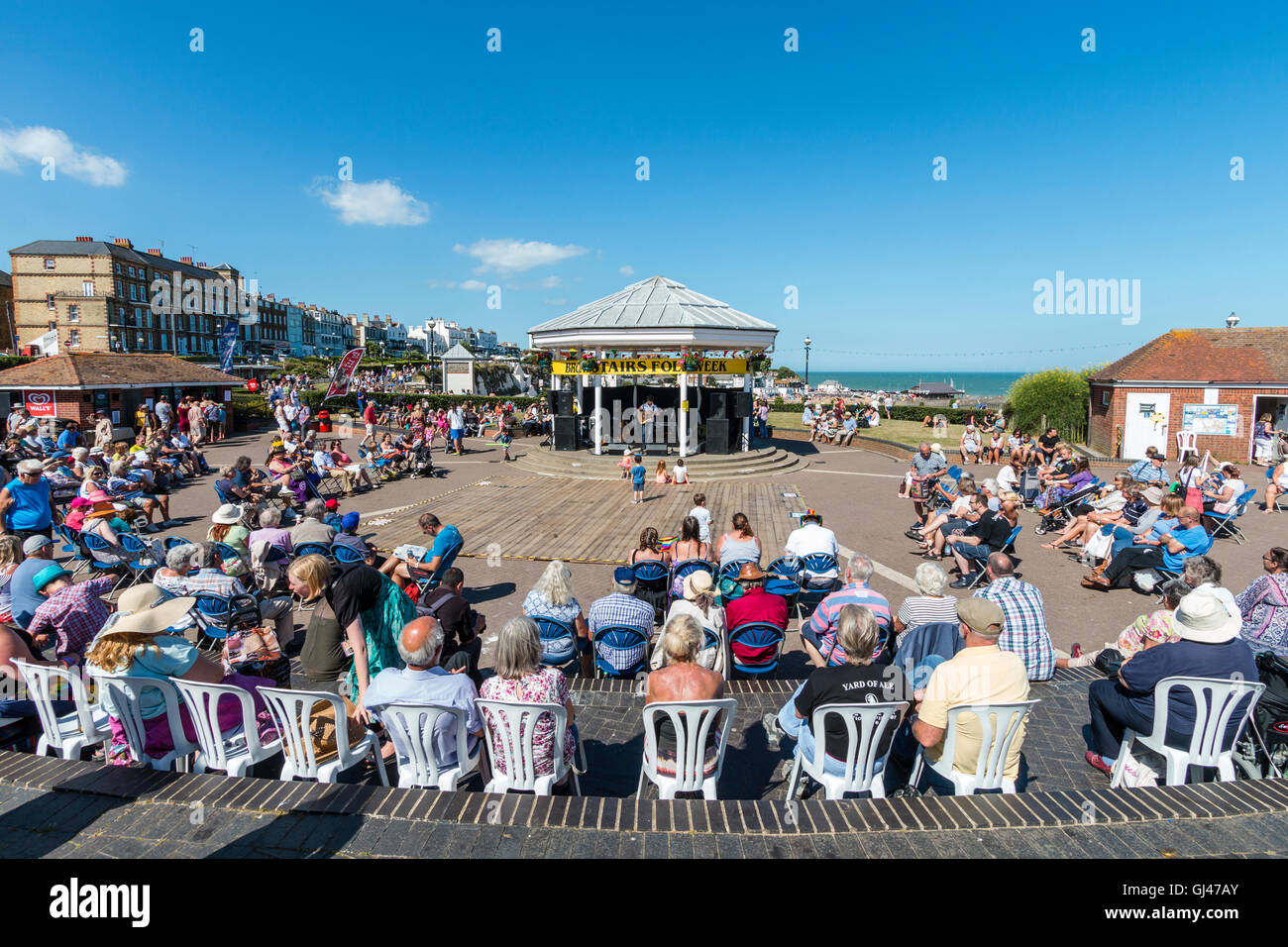 Audience of people sitting around deck and bandstand in bright sunshine ...