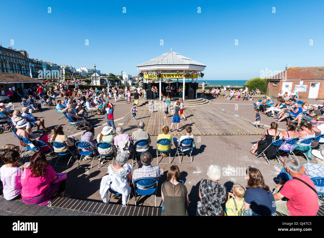 Audience of people sitting around deck and bandstand in bright sunshine ...