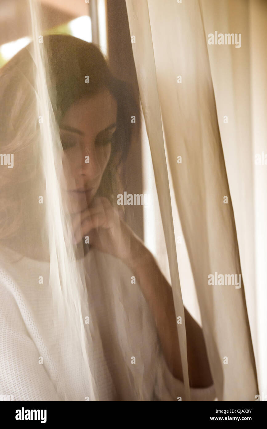 A beautiful young woman standing in the window behind the curtain and ...
