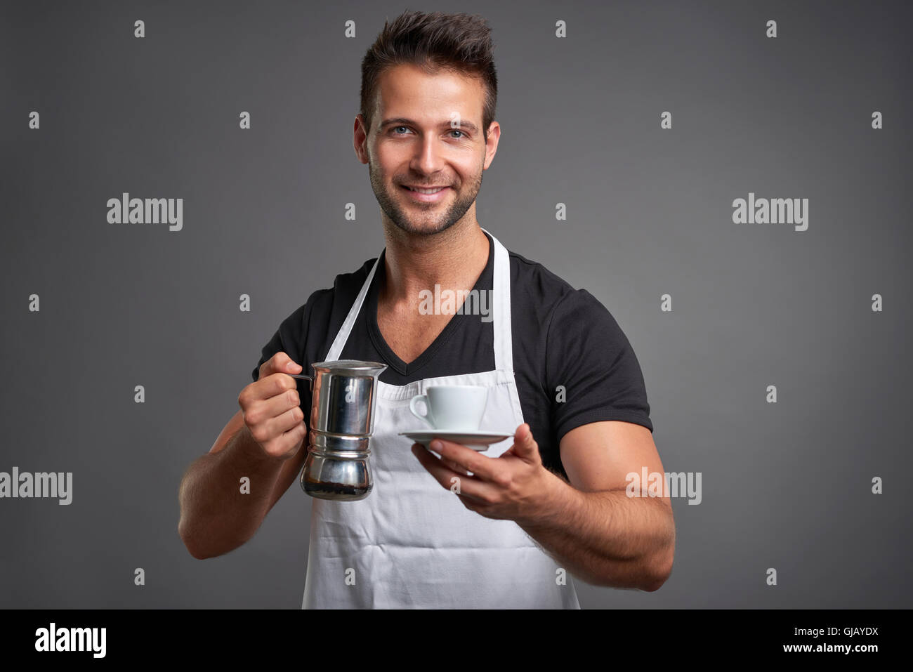 A young barista man smiling and holding a percolator and offering a cup ...