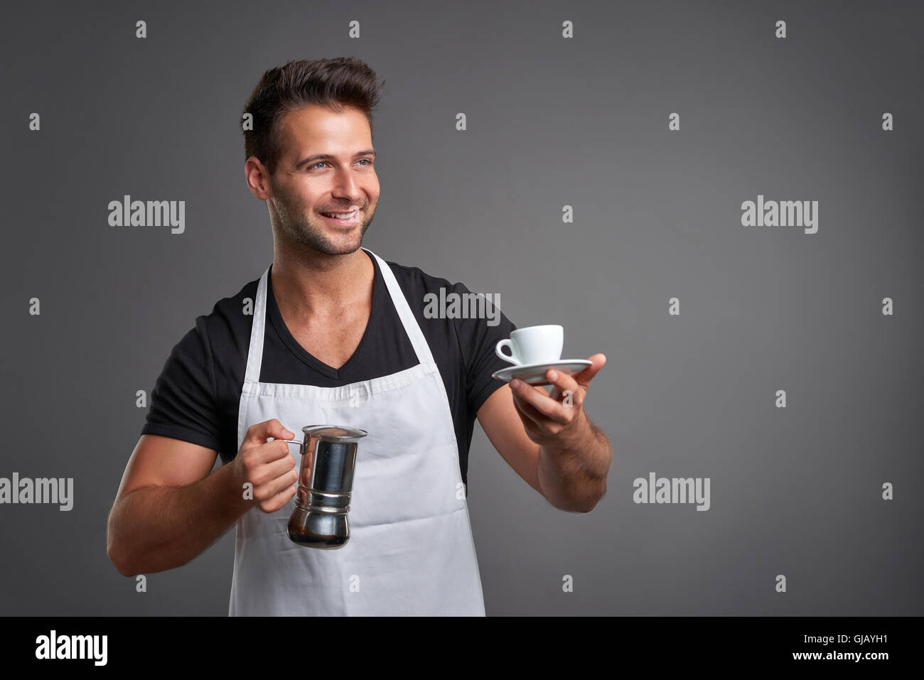 A young barista man smiling and holding a percolator and offering a cup ...