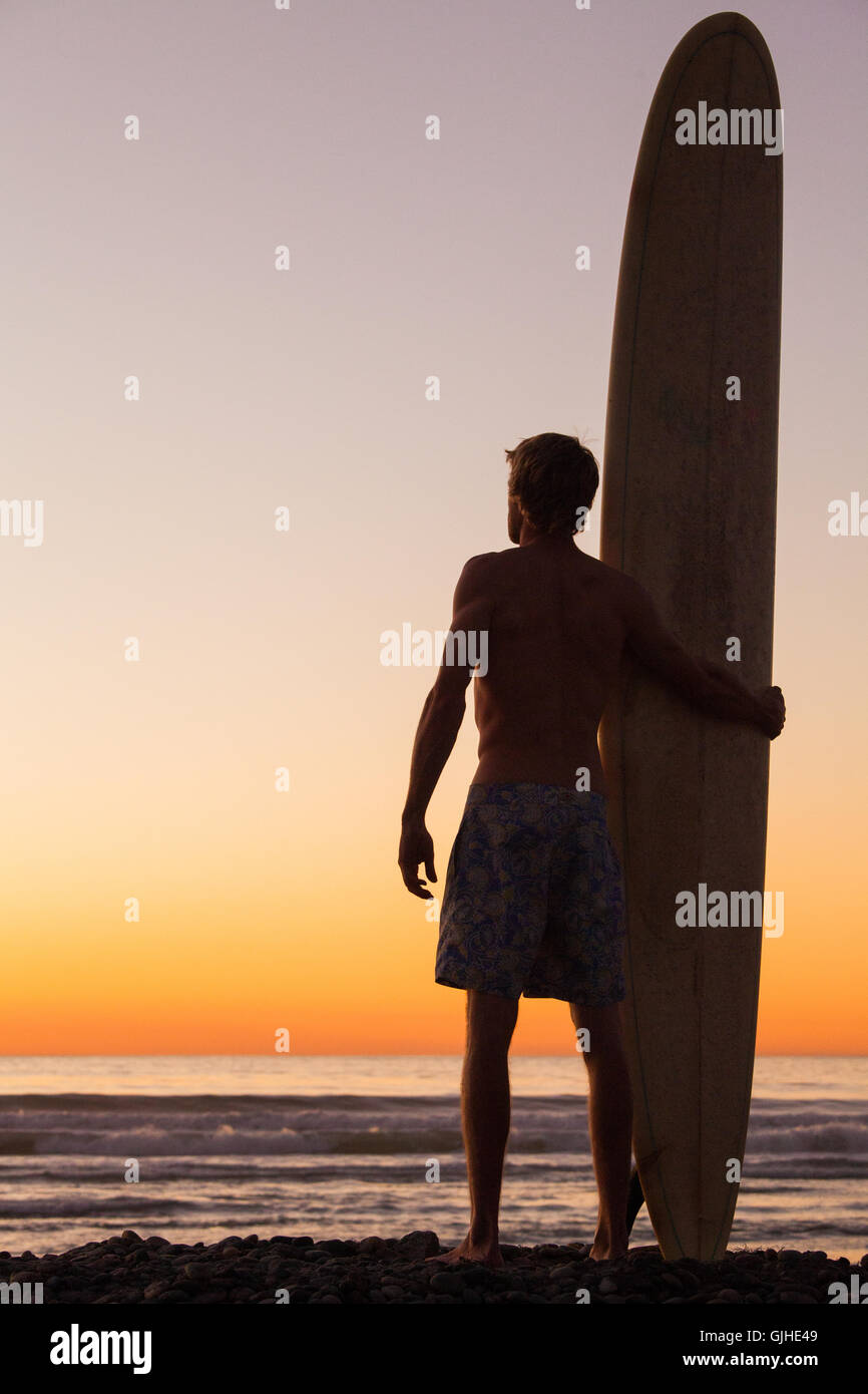 Silhouette of Man standing on beach at sunset holding longboard, San ...