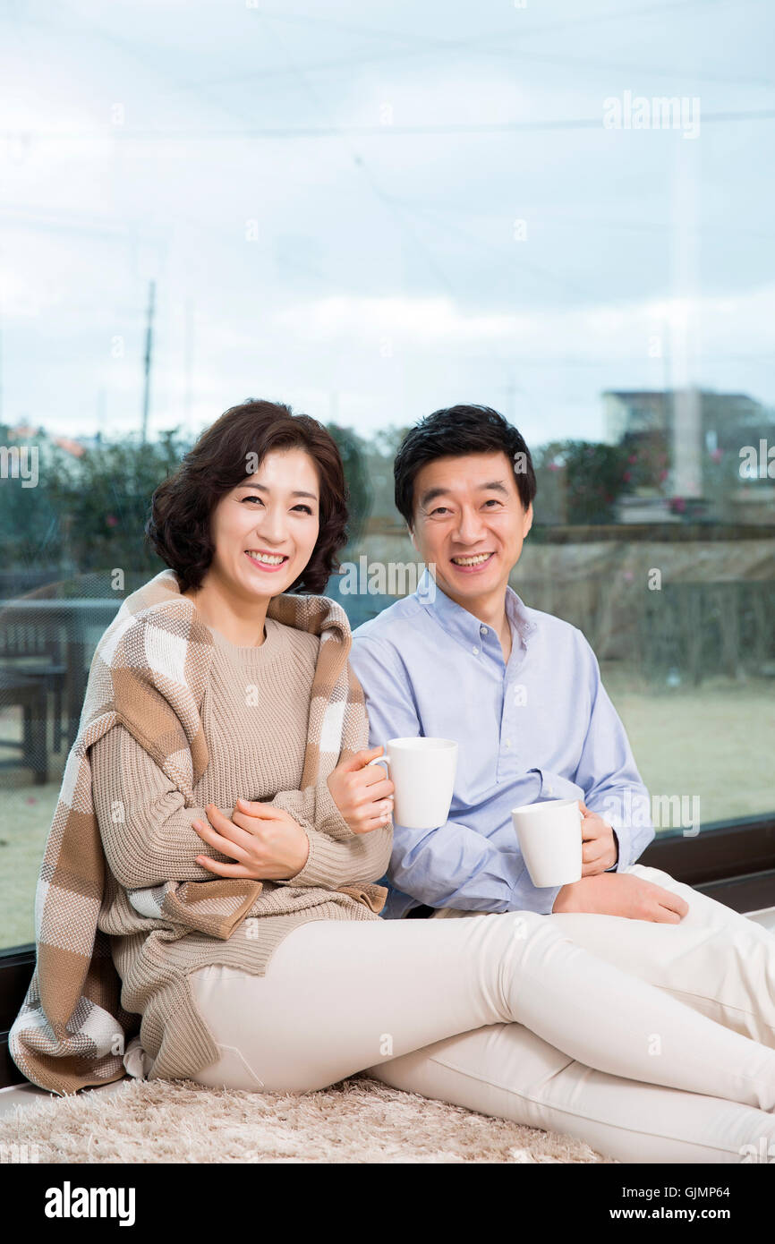 Middleaged Asian Couple Smiling And Sitting With Coffee Mugs On Rug In