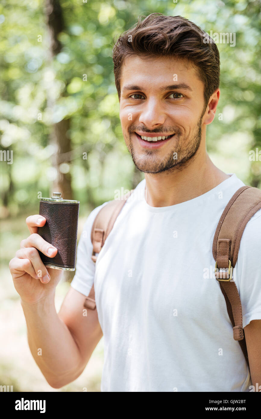 Cheerful young man with backpack standing and holding flask in forest ...