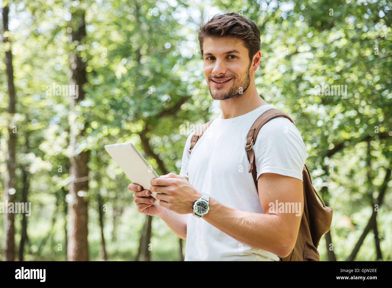 Portrait of cheerful young man with backpack using tablet in forest ...