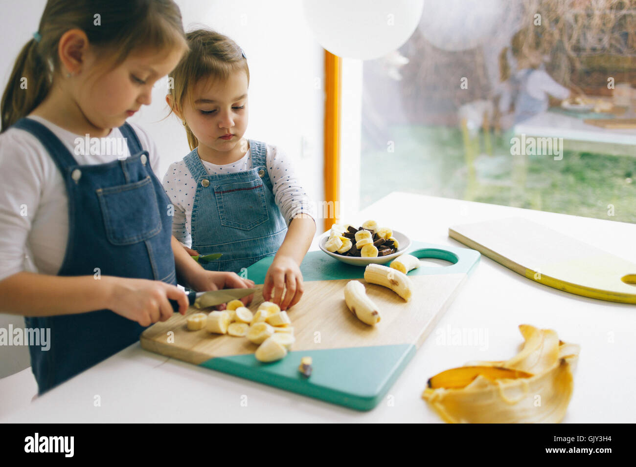 Two little girls chopping banana on the wooden board on the wooden ...