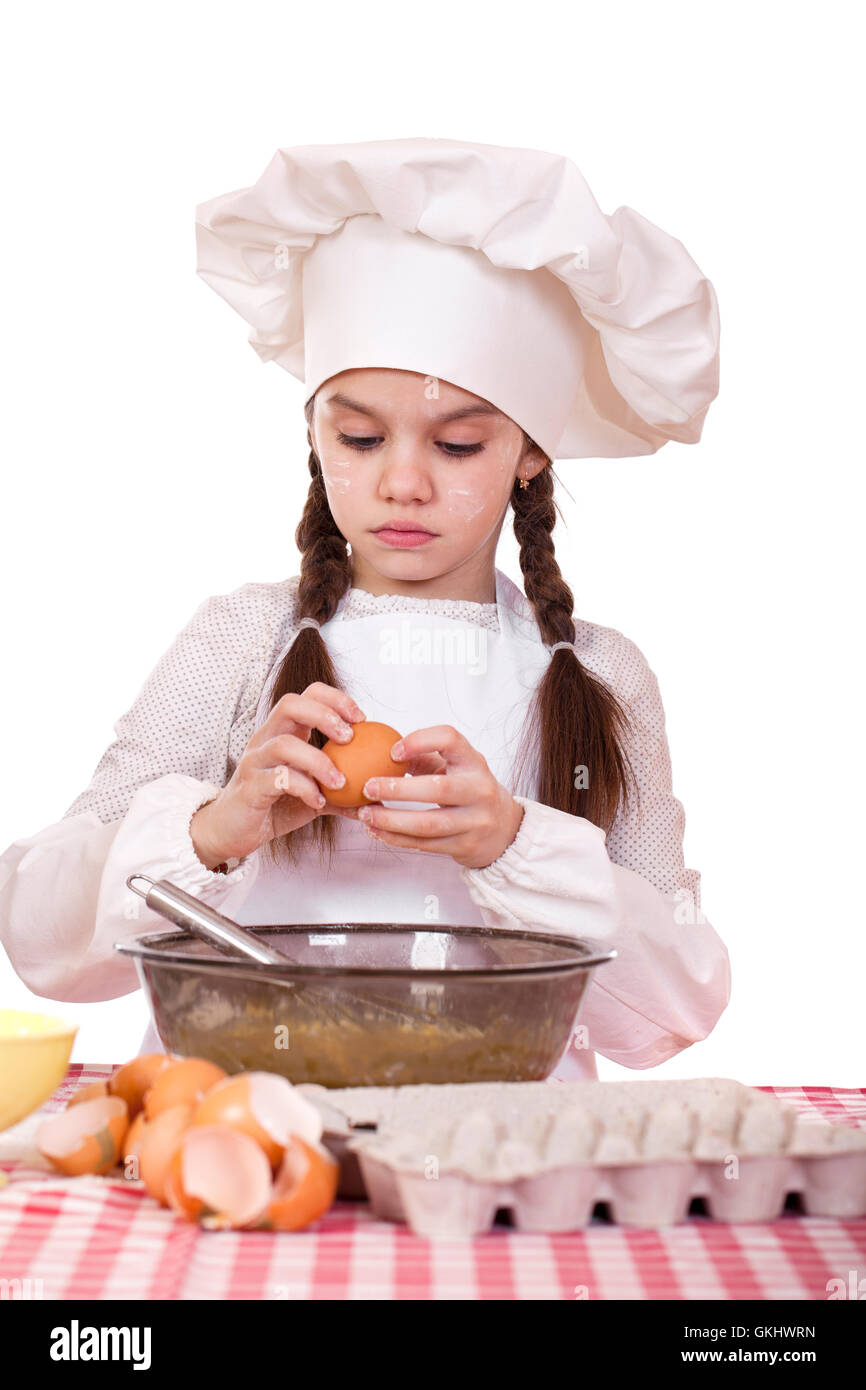 Little cook girl in a white apron breaks eggs in a deep dish, isolated ...