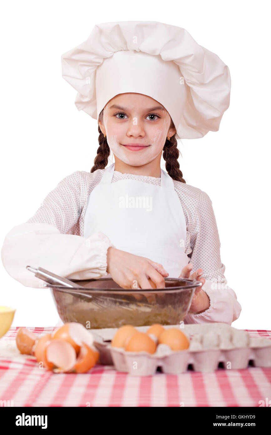 Little cook girl in a white apron breaks eggs in a deep dish, isolated ...