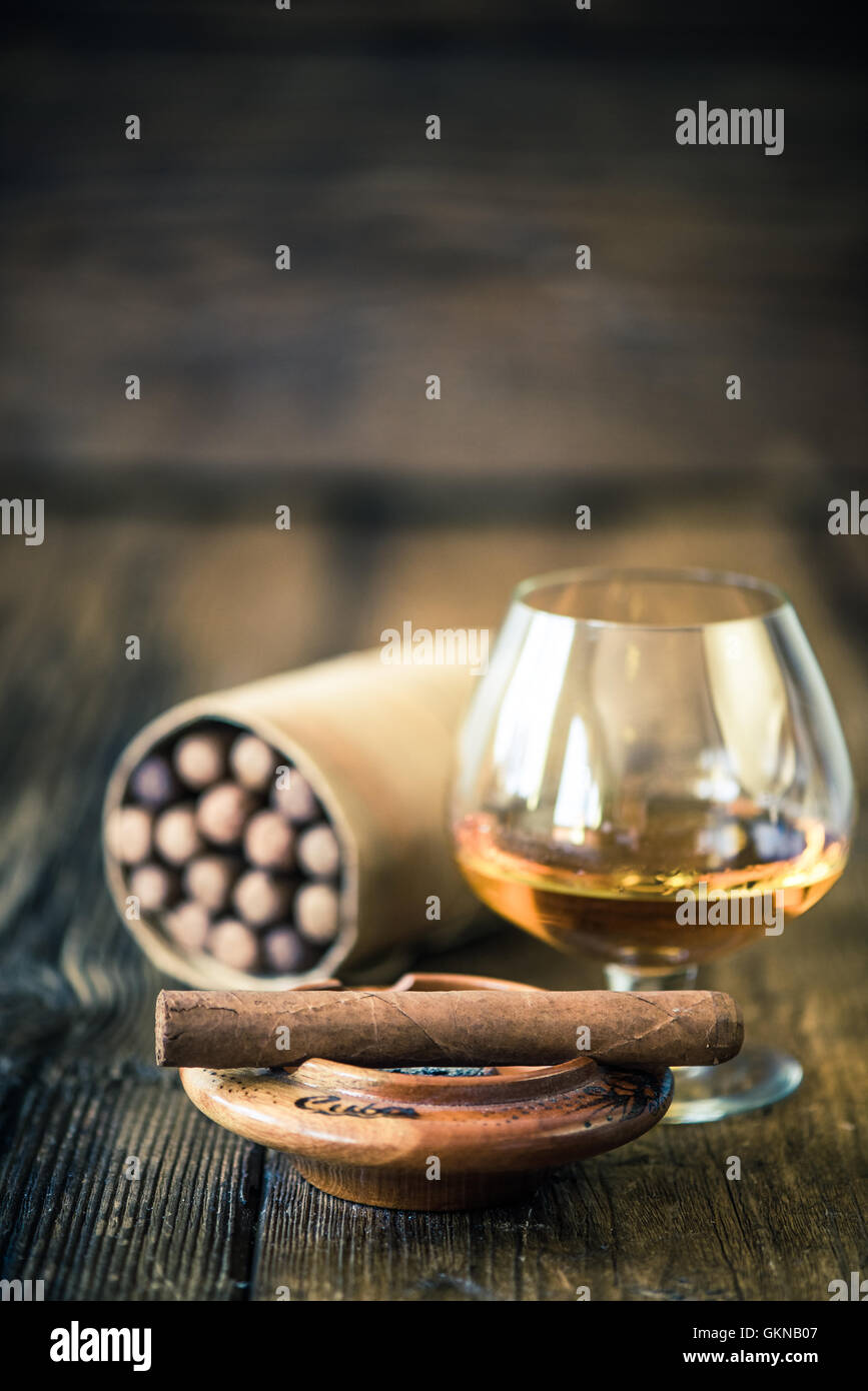 cuban cigar in ashtray on wooden table with glass of cognac or rum ...