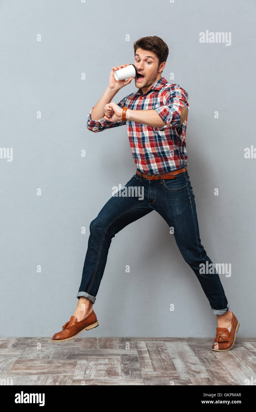 Shocked young man drinking takeaway coffee and running over grey ...