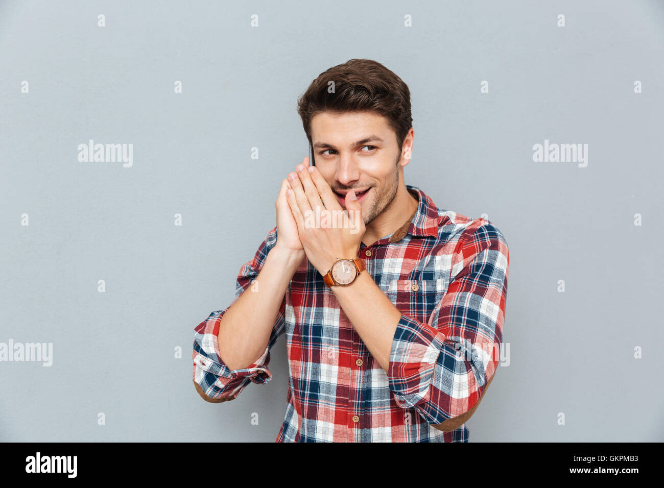 Smiling young man talking and telling secrets on cell phone over grey ...
