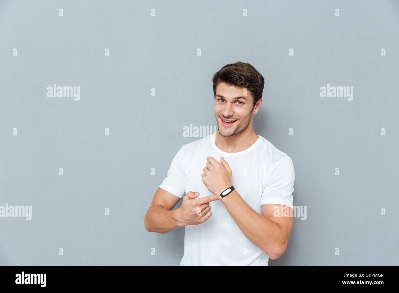 Cheerful young man standing and pointing on fitness tracker over grey ...