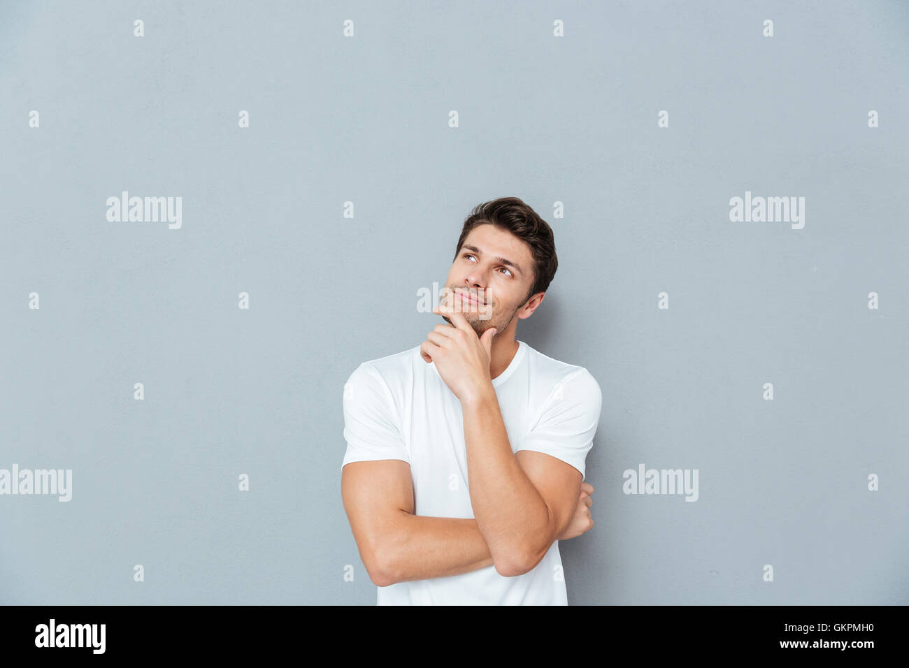 Pensive attractive young man smiling and thinking over grey background ...