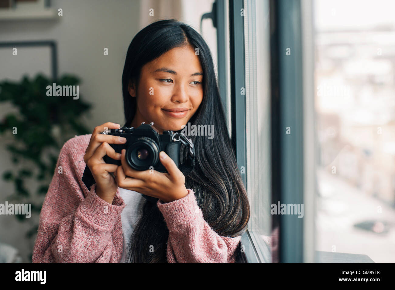 Portrait of smiling young woman with camera looking through window ...