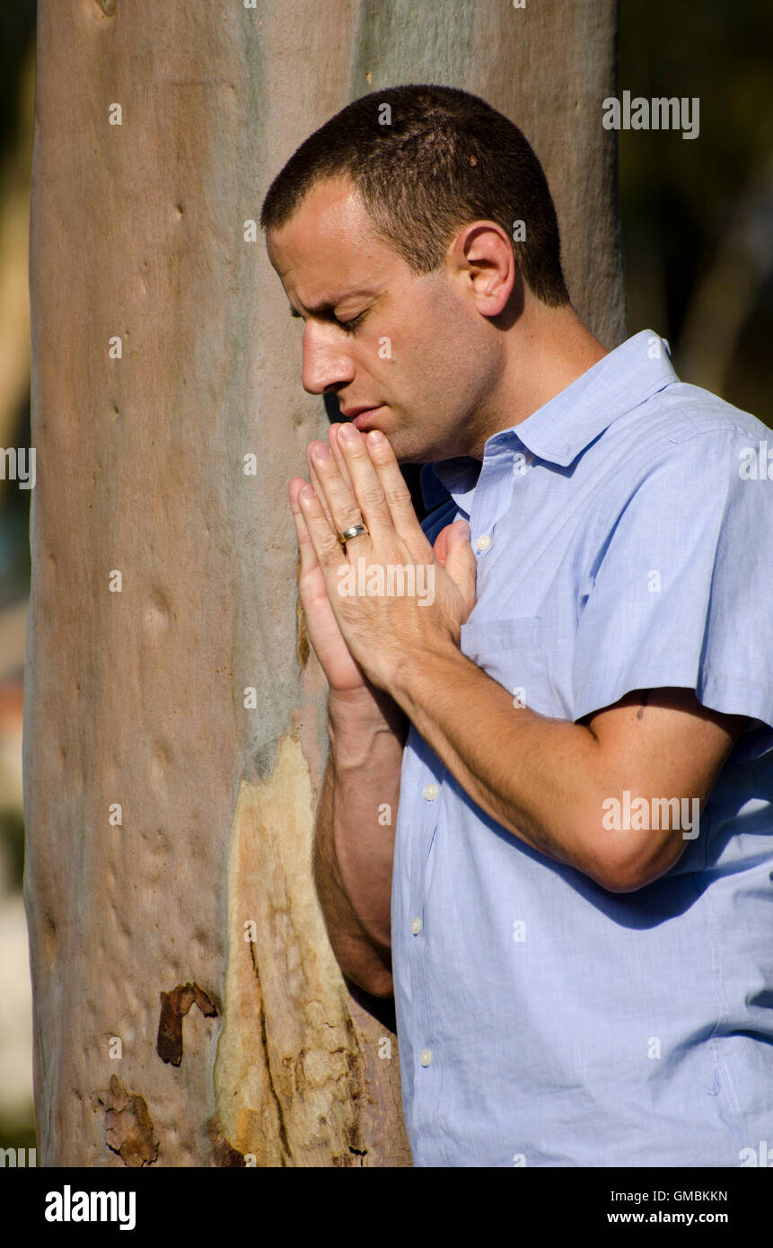 Man praying by a tree outdoors in a park wearing a blue collard shirt ...