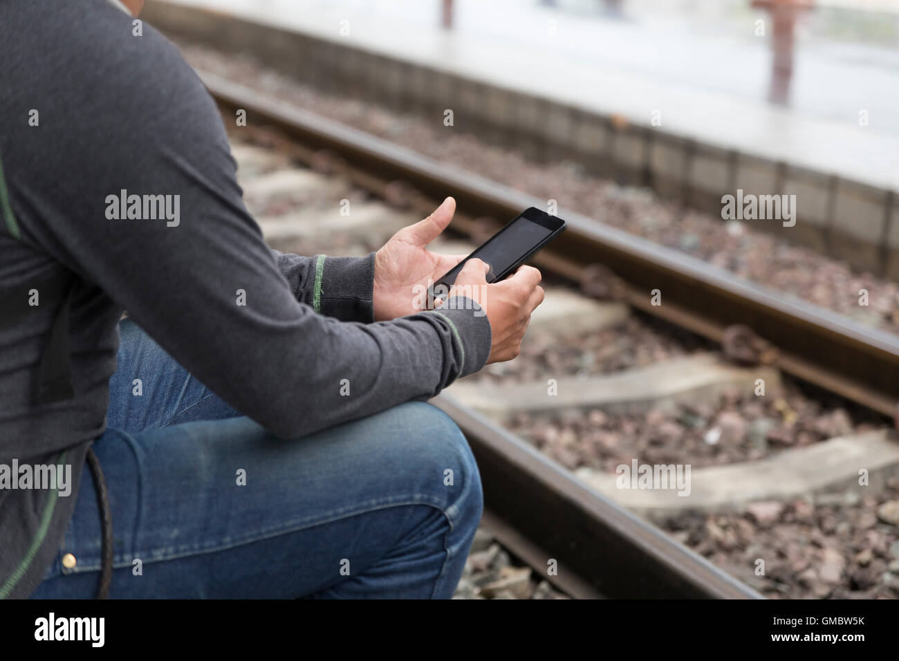 young man holding mobile phone sitting on platform at train station ...