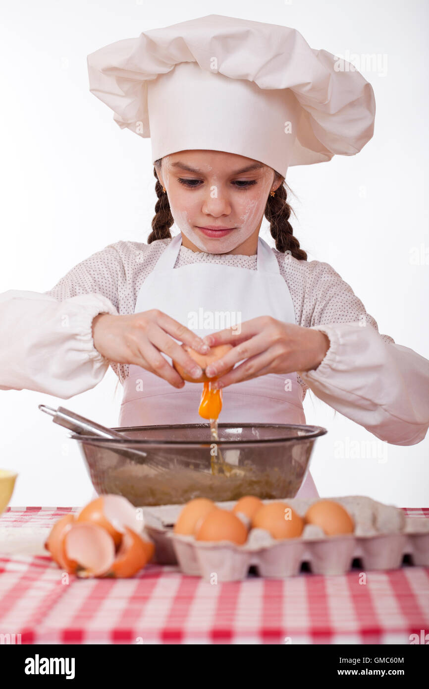 Little cook girl in a white apron breaks eggs in a deep dish, isolated ...