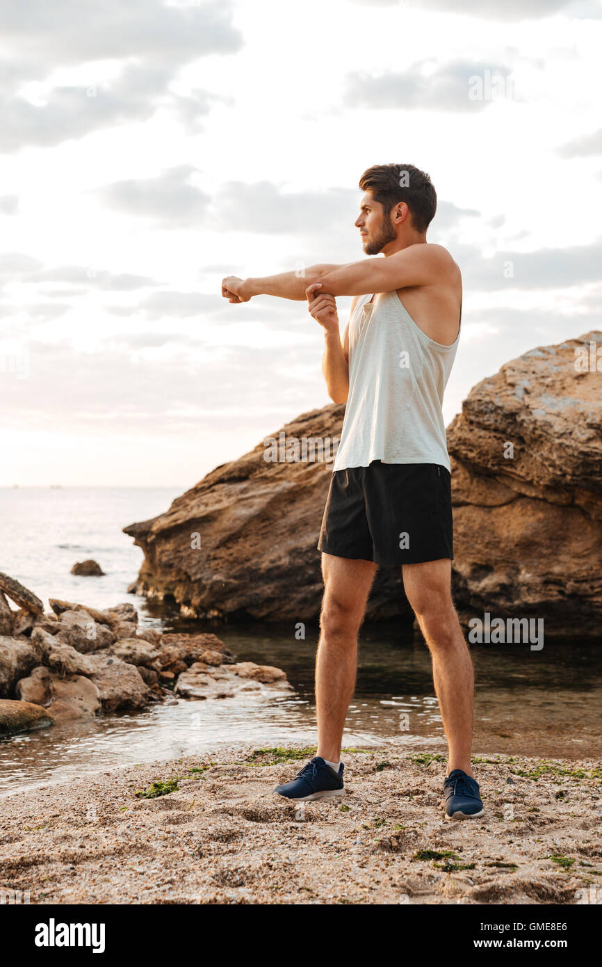 Young handsome sports man stretching hands before run at the beach ...