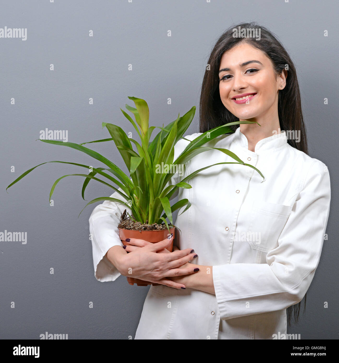 Portrait of young botanist woman in uniform holding plant against gray ...