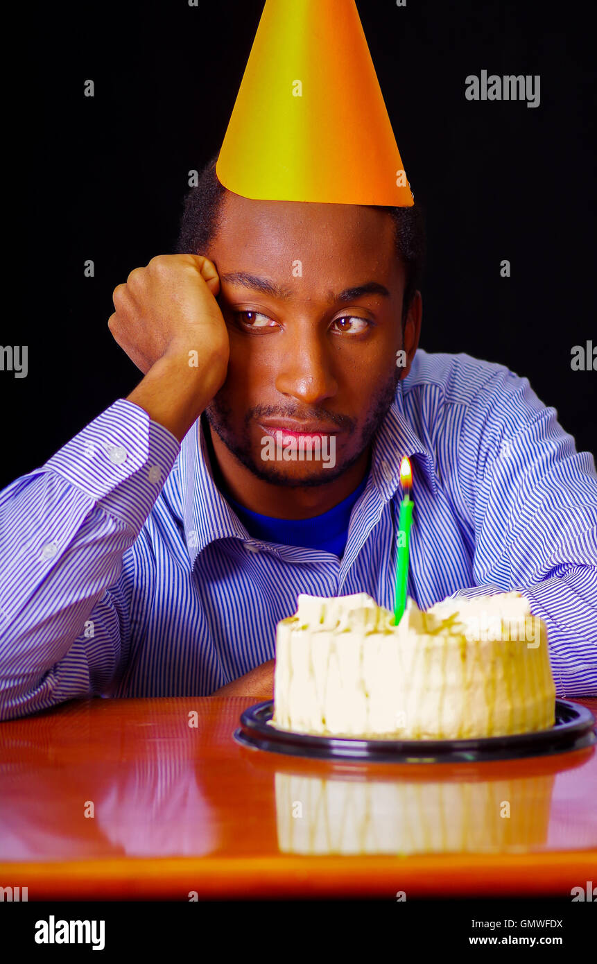 Sad man wearing blue shirt and hat sitting by table with cake in front ...