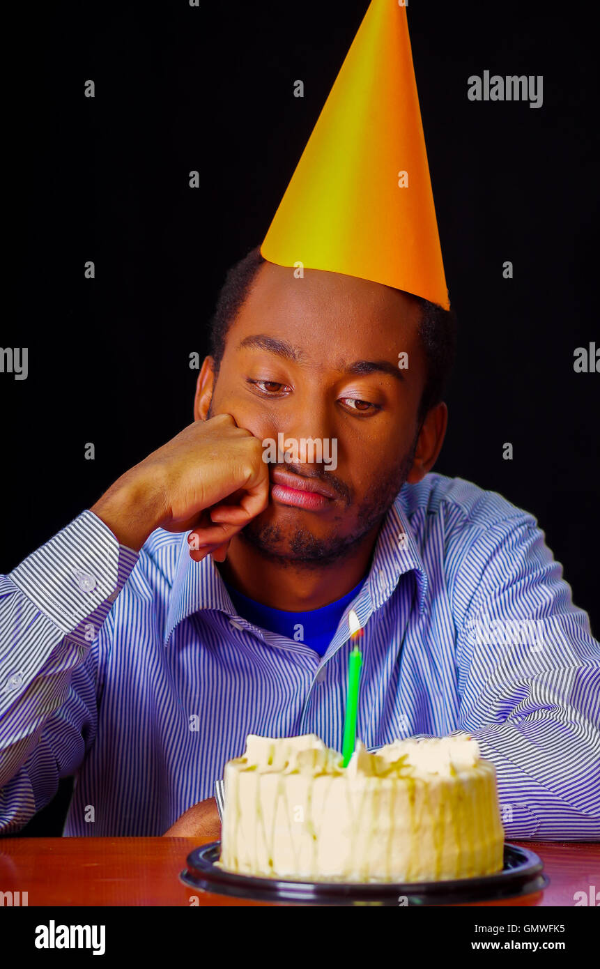 Sad man wearing blue shirt and hat sitting by table with cake in front ...