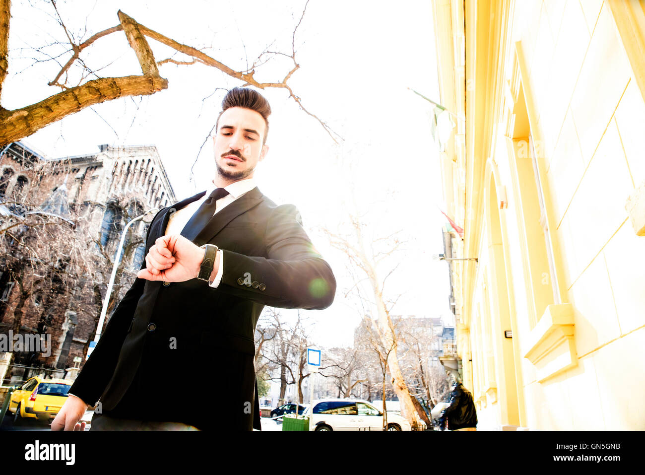 Young man walking on the street, checking the time on his wristwatch ...