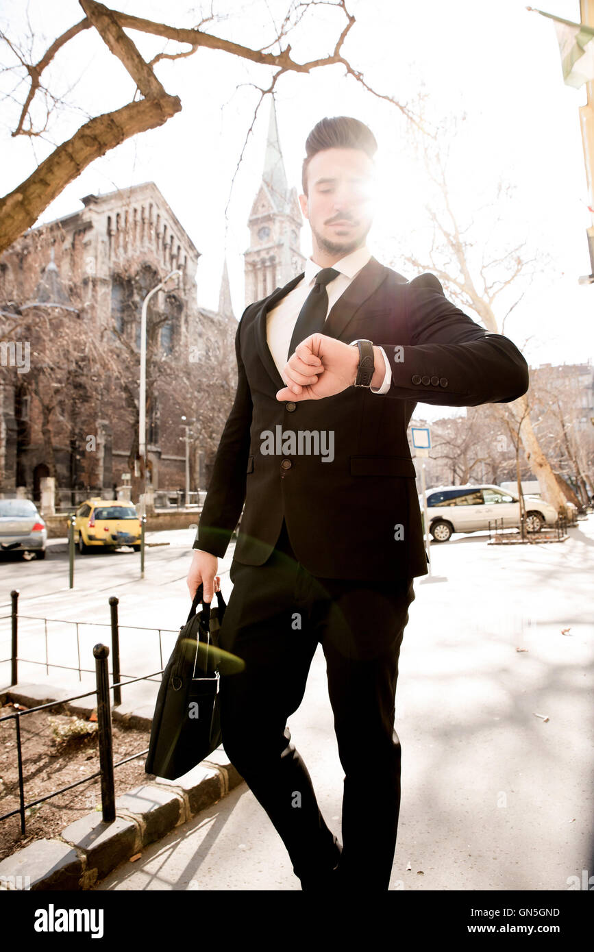 Young man walking on the street, checking the time on his wristwatch ...